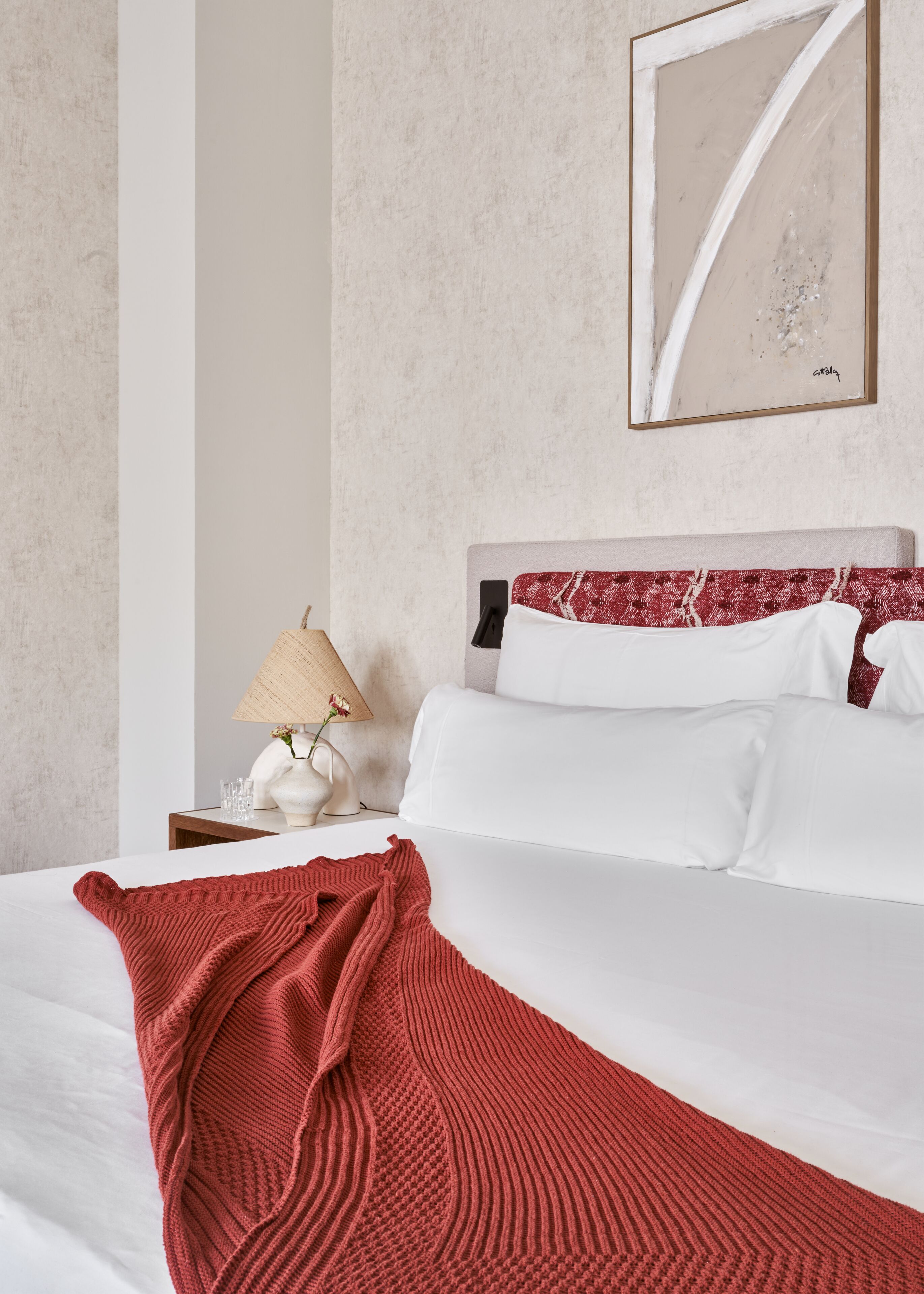Cama bem arrumada com lençóis brancos, um cobertor de tricô vermelho e uma luminária em uma mesa de cabeceira.