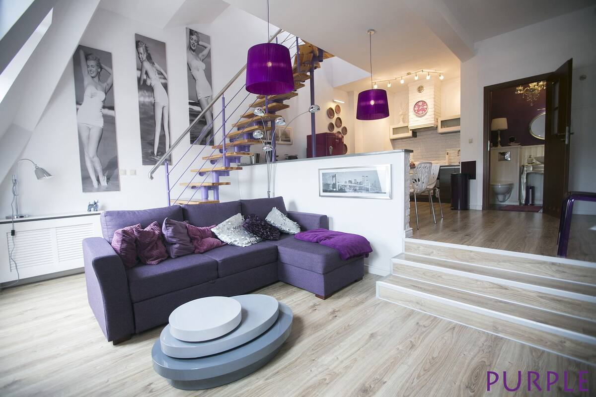 Uma sala de estar moderna com um sofá seccional roxo, uma escada e uma área de cozinha.