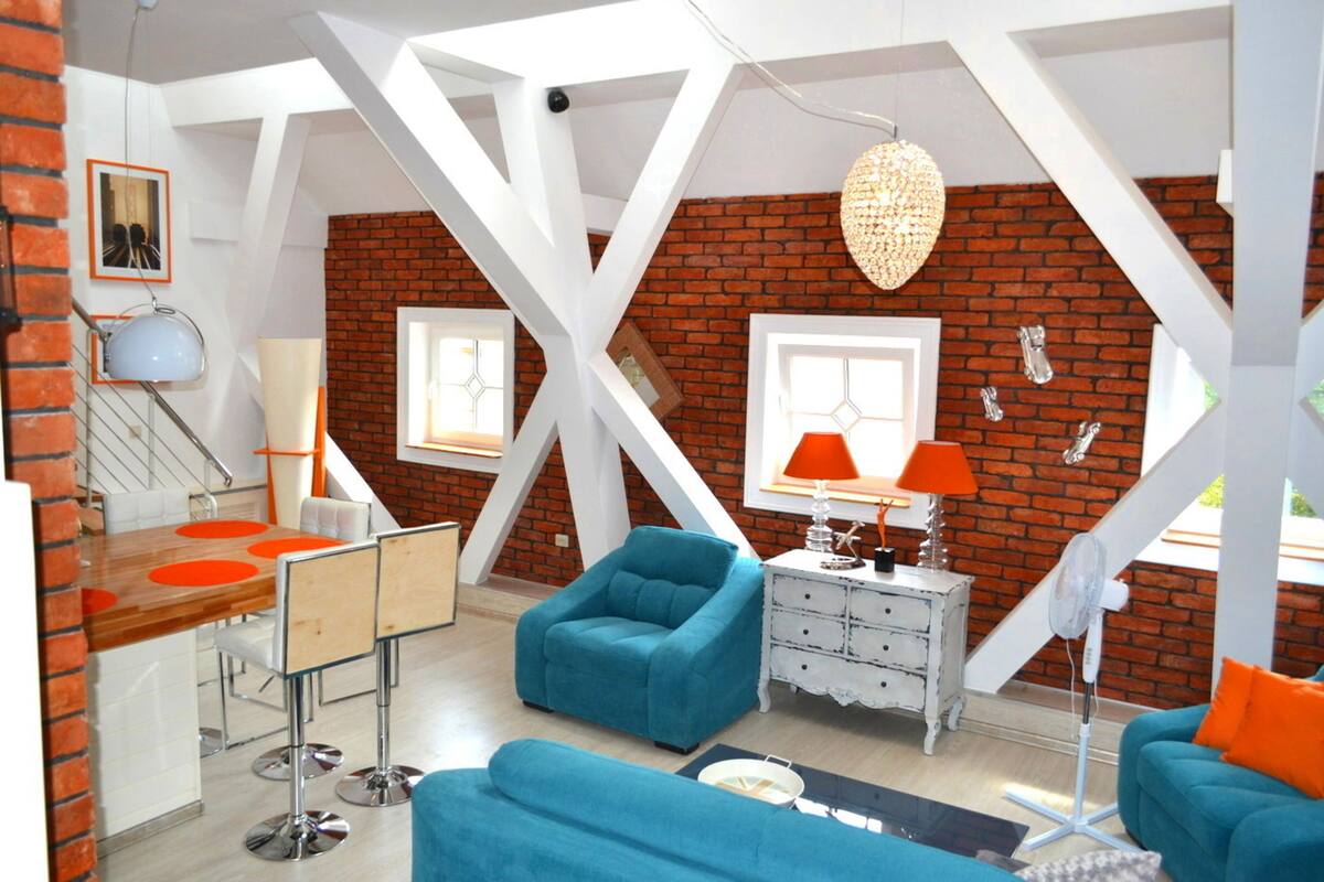 Sala moderna com parede de tijolo, vigas brancas, área de jantar com cadeiras laranja e um sofá azul.