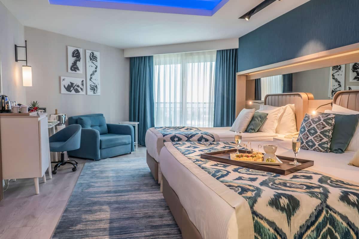 Quarto de hotel com duas camas, uma escrivaninha, uma cadeira e uma janela grande com cortinas.