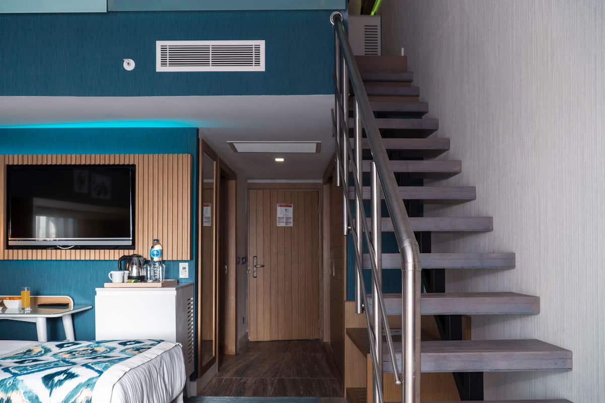 Quarto de hotel com cama, escrivaninha, televisão, escada e porta.