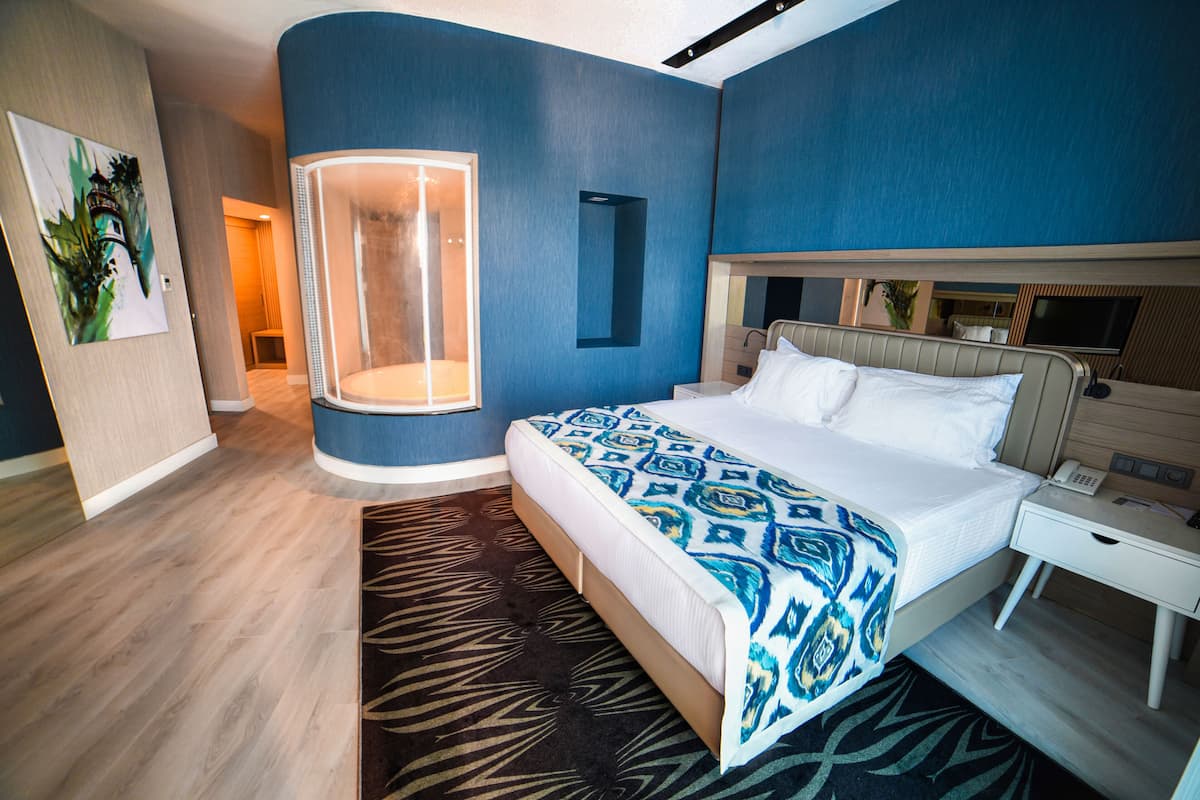 Quarto de hotel moderno com uma cama grande, área de estar com sofás curvos e uma pequena mesa de cabeceira com telefone.