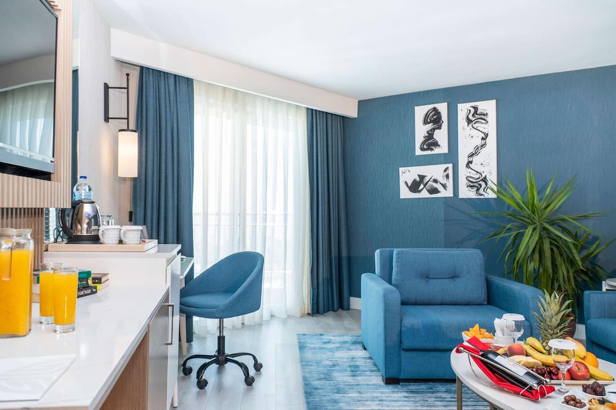 Quarto de hotel moderno com área de jantar, um sofá azul, uma mesa de centro com frutas e uma garrafa, e obras de arte na parede.