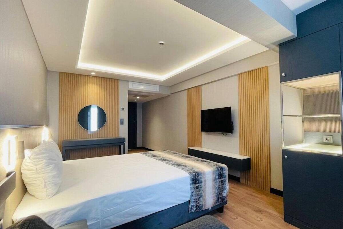 Quarto de hotel moderno com cama grande, televisão de tela plana, armário embutido e mesa de cabeceira.