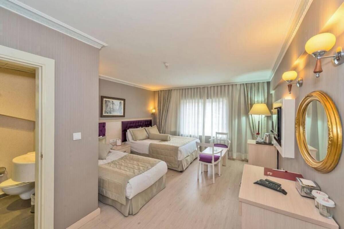 Quarto de hotel com duas camas, uma escrivaninha, uma cadeira, um espelho e uma janela com cortinas.