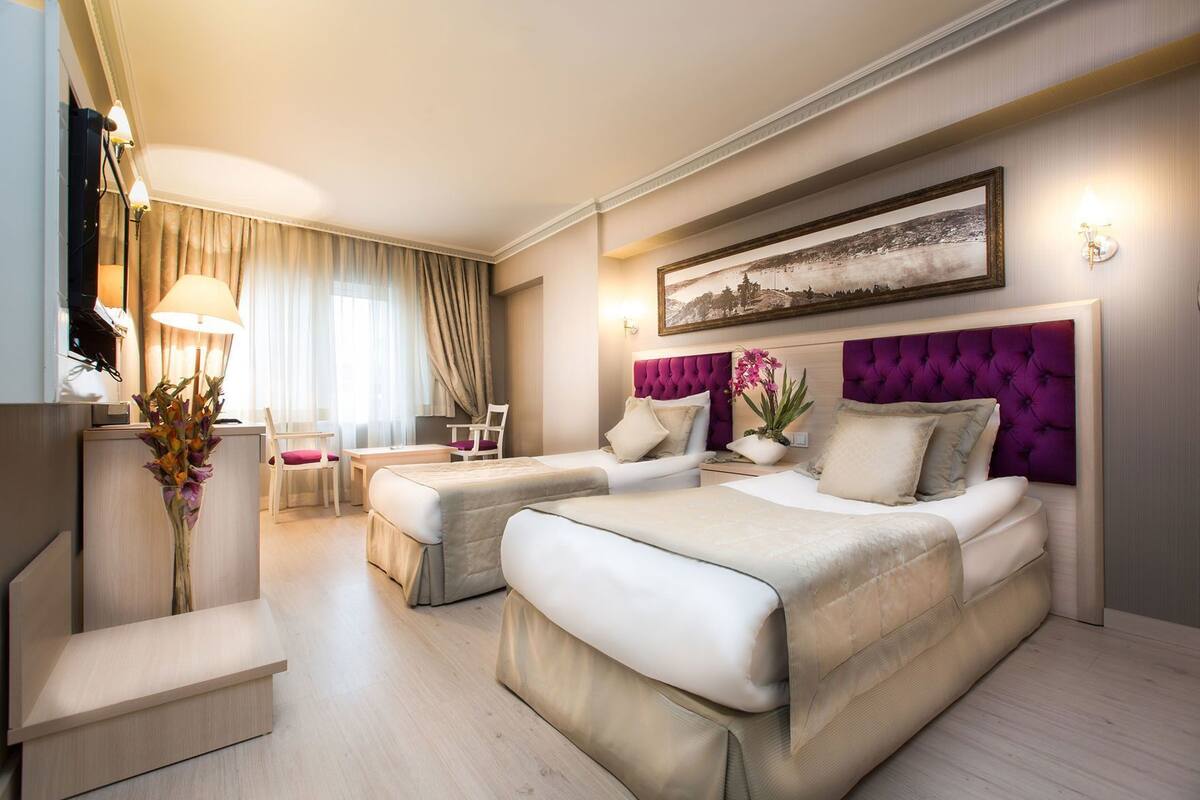 Quarto de hotel com duas camas, uma mesa, uma cadeira, luminária de mesa, um vaso com flores e um quadro na parede.