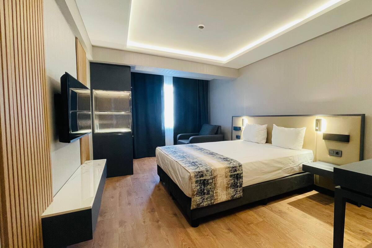 Quarto de hotel moderno com uma cama grande, escrivaninha, cadeira e televisão na parede.