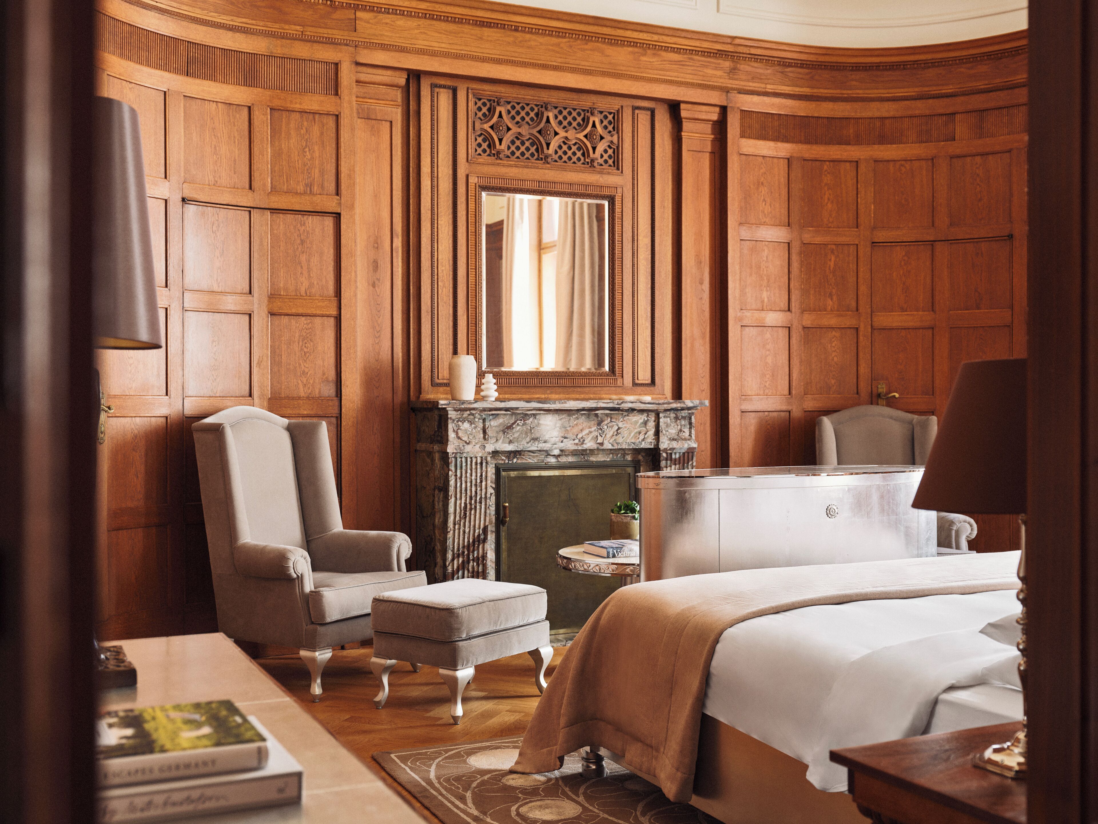 Quarto de hotel com uma cama grande, lareira, área de estar com uma poltrona e um banquinho, e lambris de madeira nas paredes.