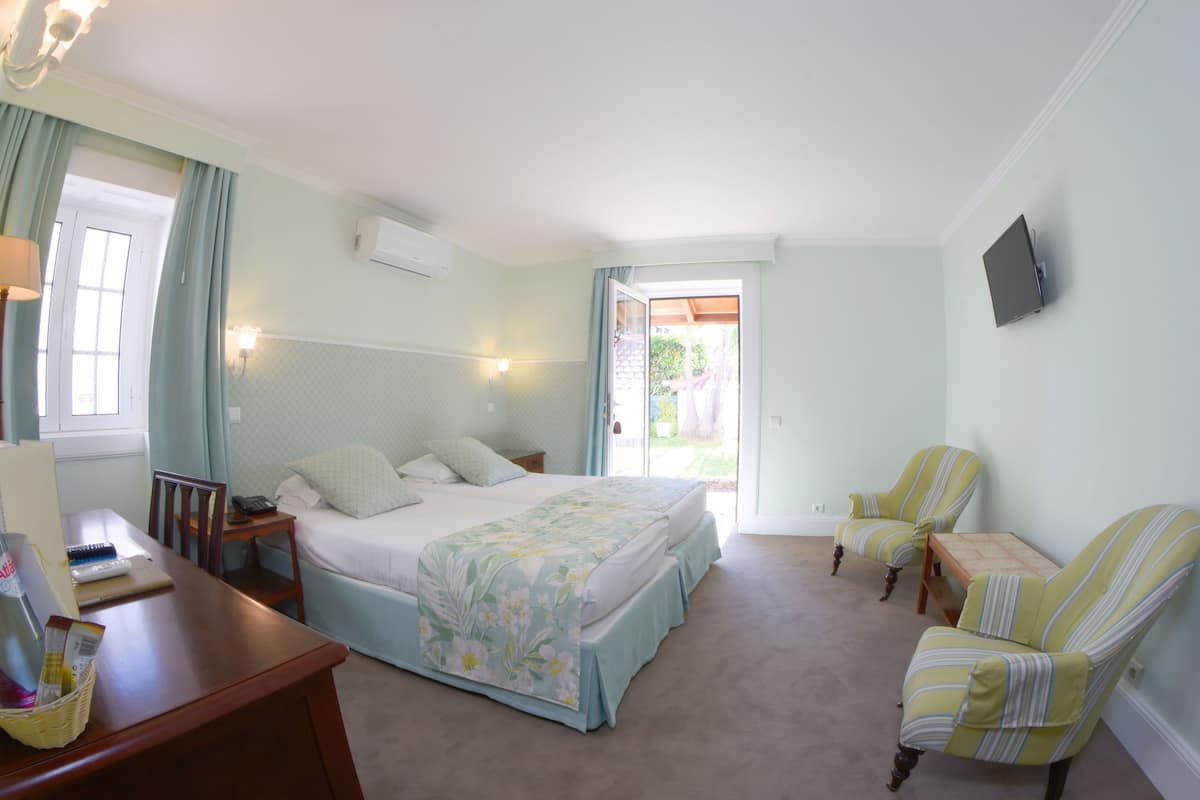 Quarto com cama, duas cadeiras, uma mesa e televisão na parede.