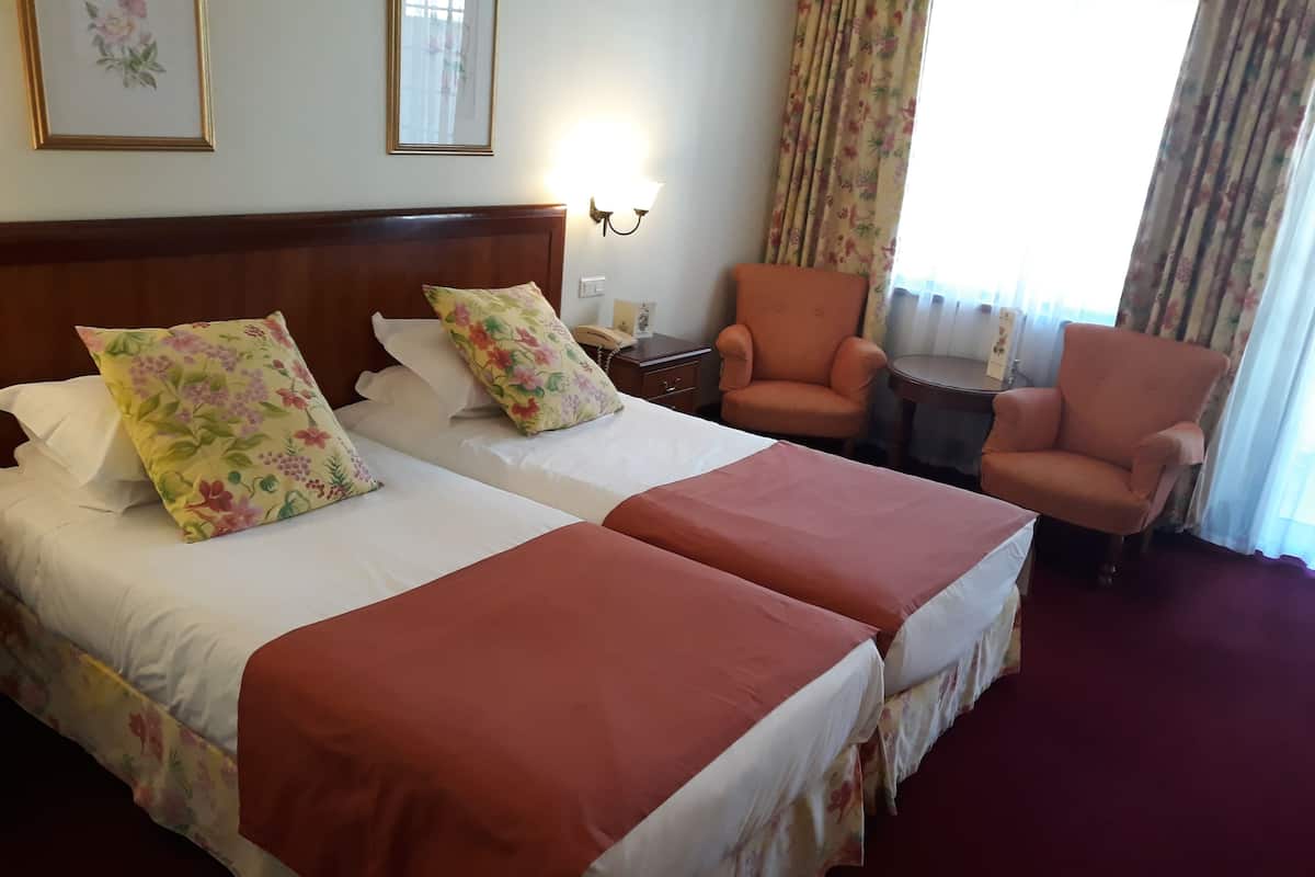 Quarto de hotel com duas camas, almofadas estampadas com padrão floral, cabeceira de madeira, uma pequena mesa, uma poltrona laranja e uma janela com cortinas.