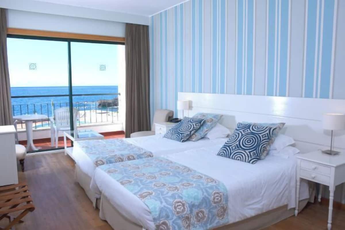 Quarto de hotel com uma cama grande, duas mesas de cabeceira, uma escrivaninha com cadeira e uma varanda com vista para o mar.