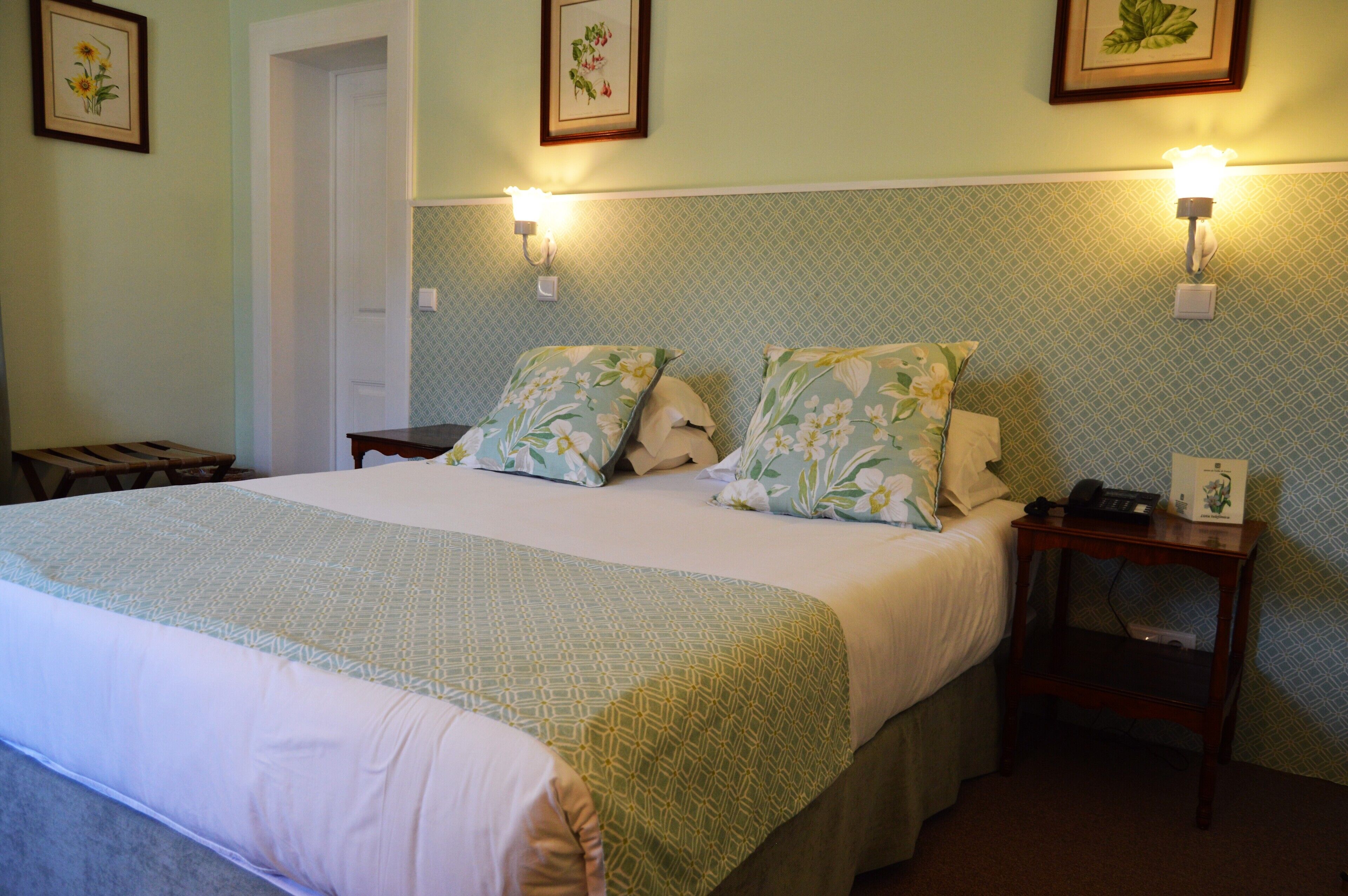 Cama bem arrumada com roupa de cama florida, mesa de cabeceira com abajur e quadro com flores na parede.