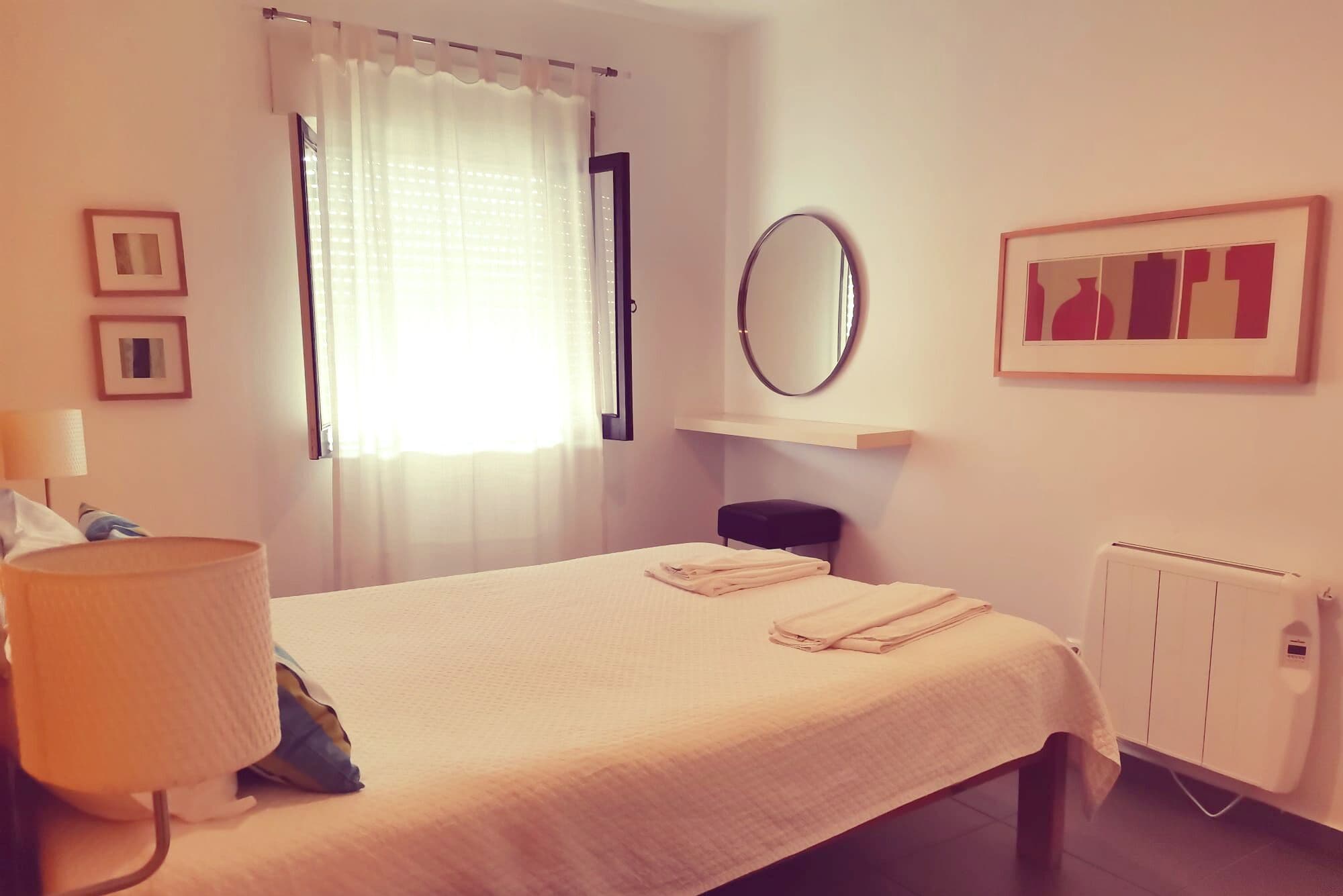 Quarto de hotel com cama, luminária de cabeceira, janela com cortinas, espelho, quadro emoldurado e radiador.