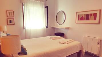 Quarto de hotel com cama, luminária de cabeceira, janela com cortinas, espelho, quadro emoldurado e radiador.