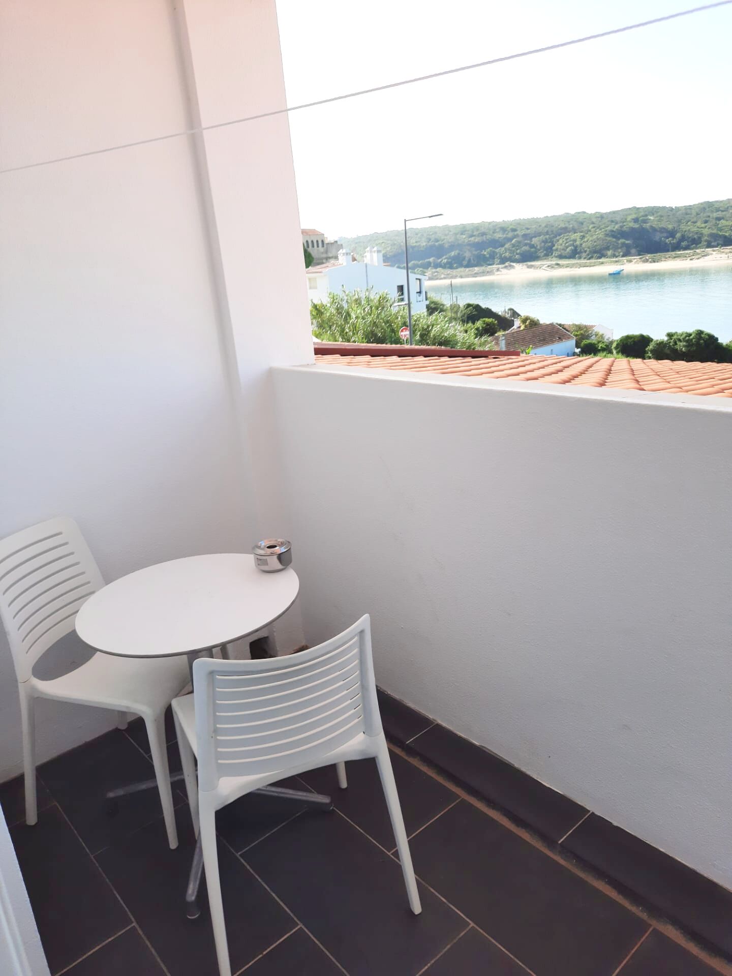 Varanda com mesa e duas cadeiras brancas, vista para área costeira com praia e corpo d'água.