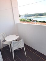 Varanda com mesa e duas cadeiras brancas, vista para área costeira com praia e corpo d'água.