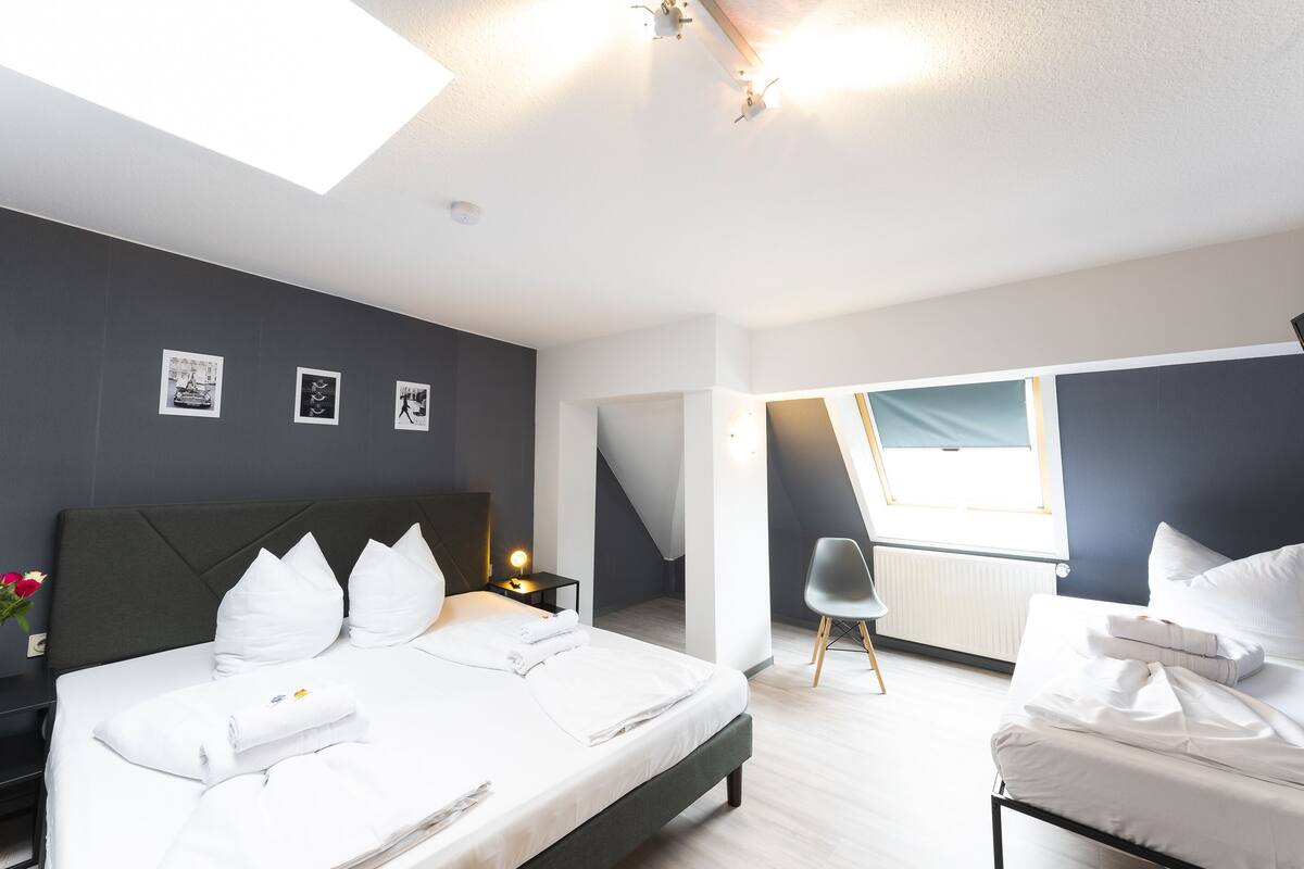 Quarto de hotel moderno com uma cama grande, uma escrivaninha e uma cadeira. Há um clarabóia acima da cama e uma janela com vista para o exterior.