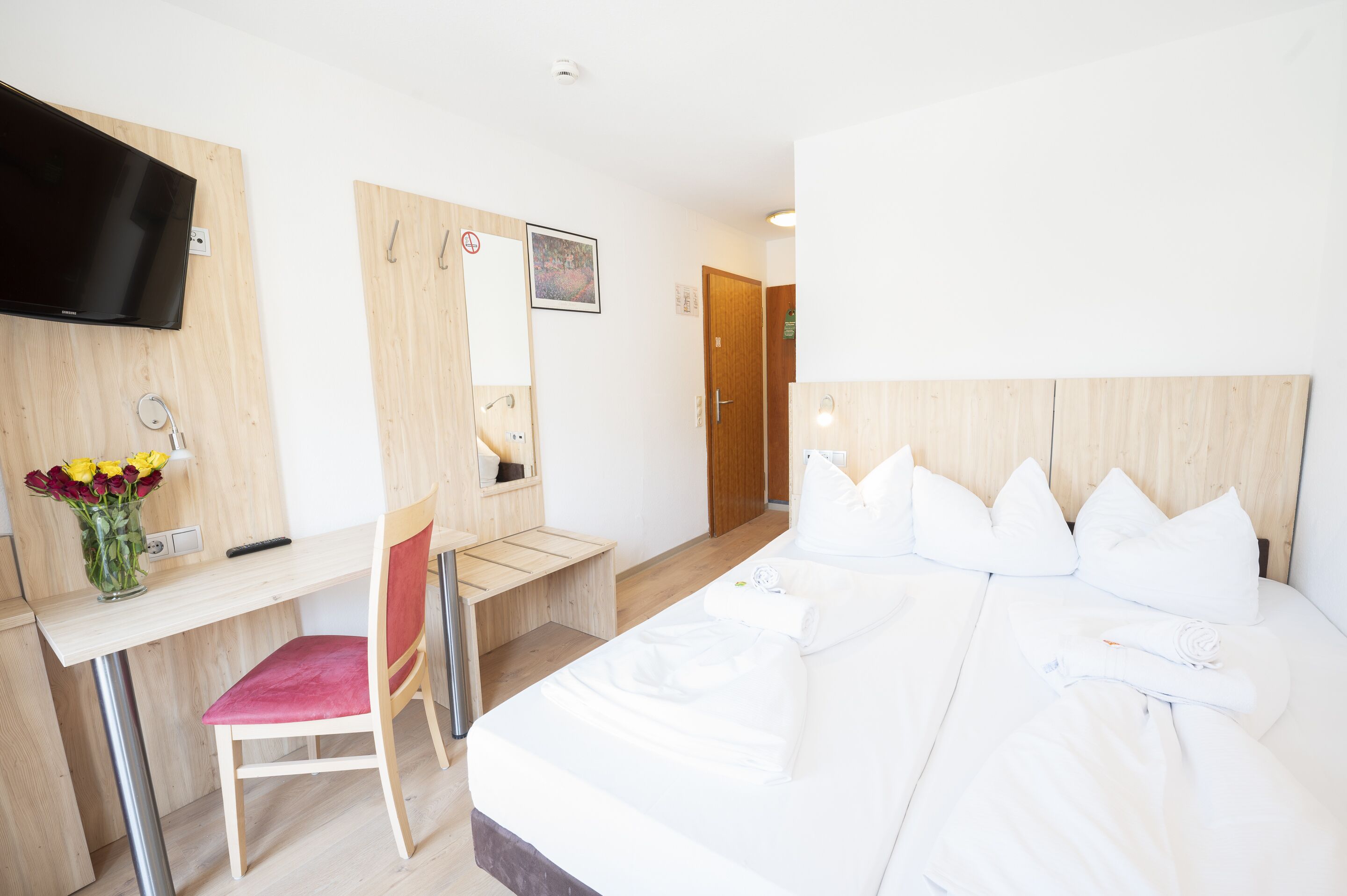Quarto de hotel com cama, escrivaninha, cadeira, televisão e porta do banheiro.
