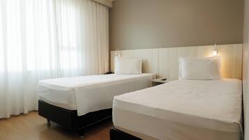 Duas camas de solteiro com lençóis brancos, piso de madeira e janela com cortinas leves.