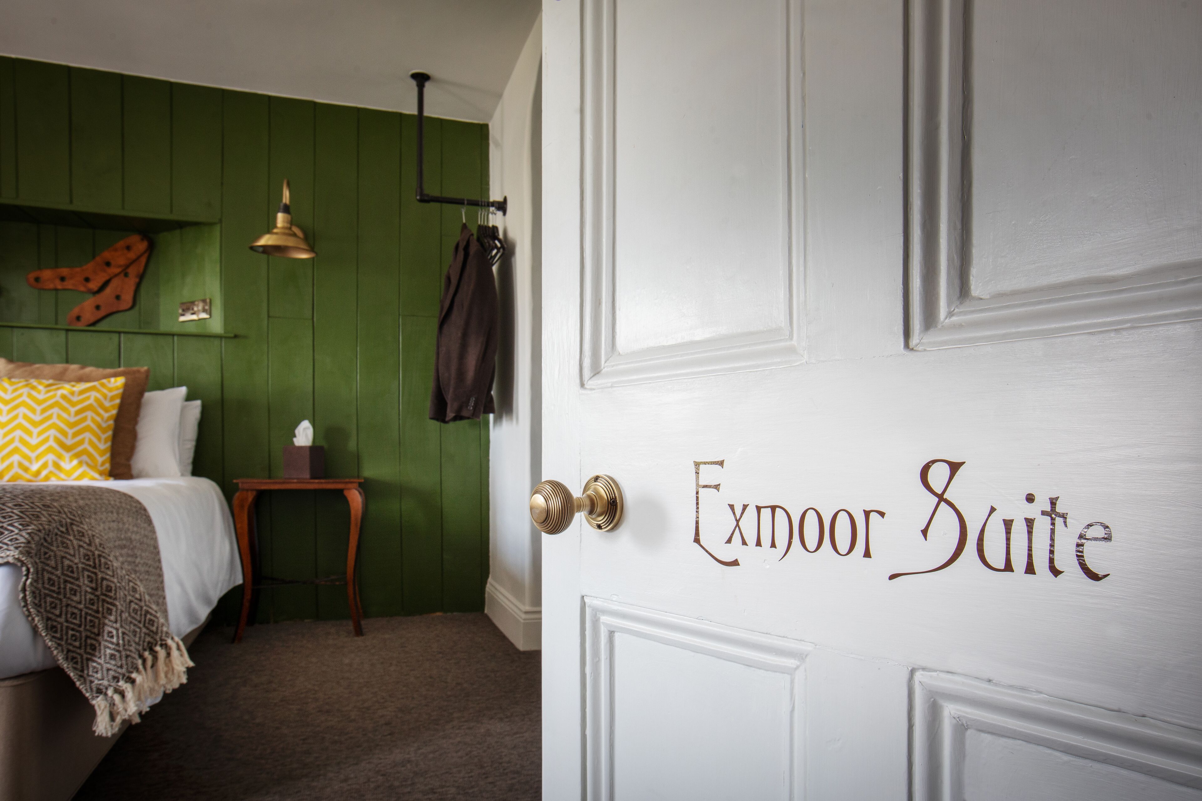 Um quarto com uma cama, uma mesinha de cabeceira com um estojo de lenços, uma parede verde e uma porta com a inscrição "Exmoor Suite".