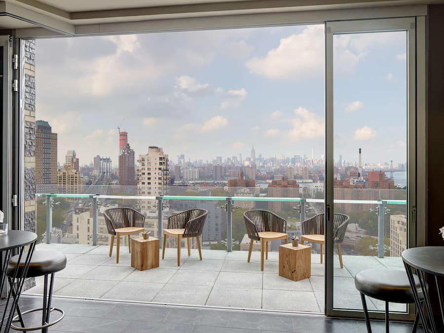 Varanda com vista para a cidade, equipada com cadeiras e mesas, e guarda-corpo de vidro.