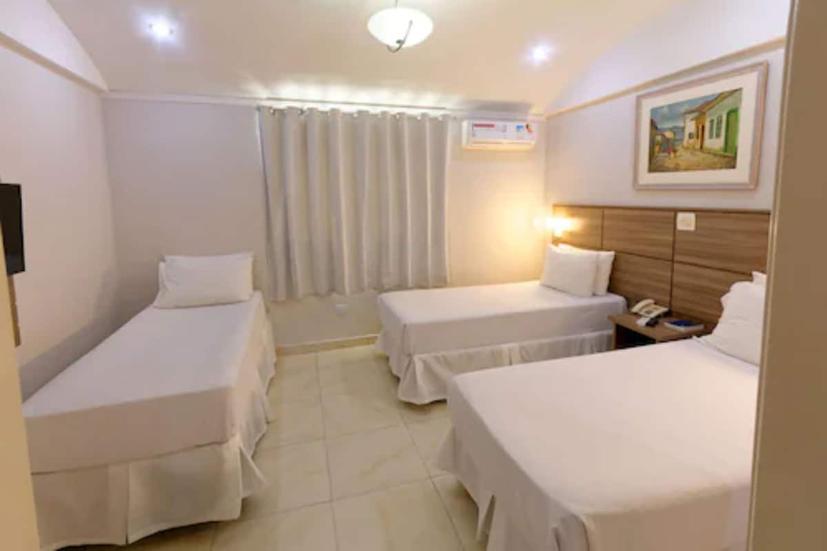 Quarto de hotel com duas camas de solteiro, uma mesa de cabeceira com luminária, um quadro na parede e uma janela com cortinas.