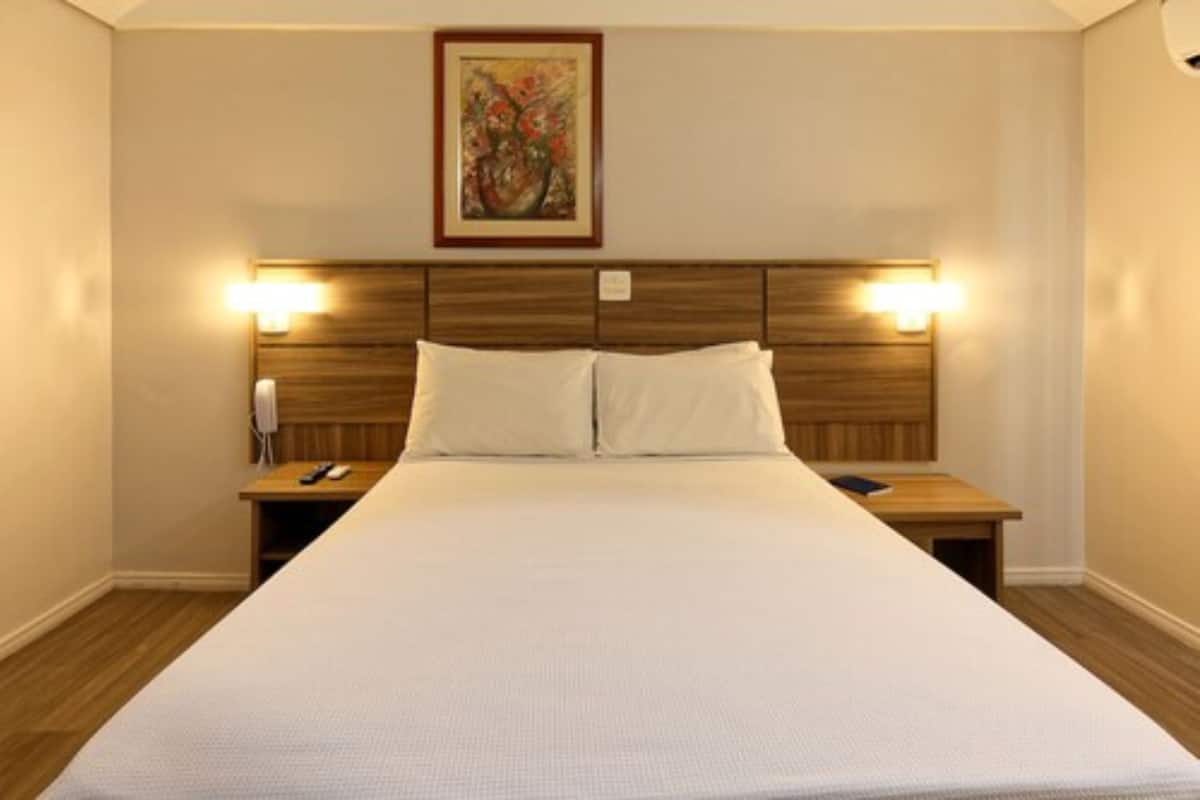 Quarto de hotel com uma cama grande, duas mesas de cabeceira, um painel de cabeceira de madeira e uma obra de arte emoldurada na parede.