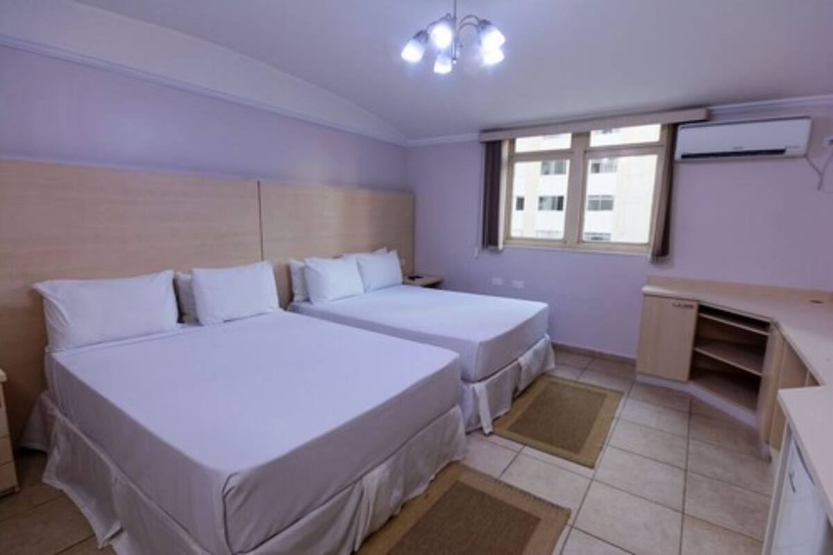Quarto de hotel com duas camas, uma escrivaninha, uma janela e ar condicionado.