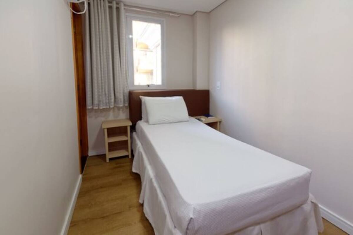 Quarto pequeno com uma cama de solteiro, cabeceira de madeira, mesa de cabeceira e janela com cortinas.
