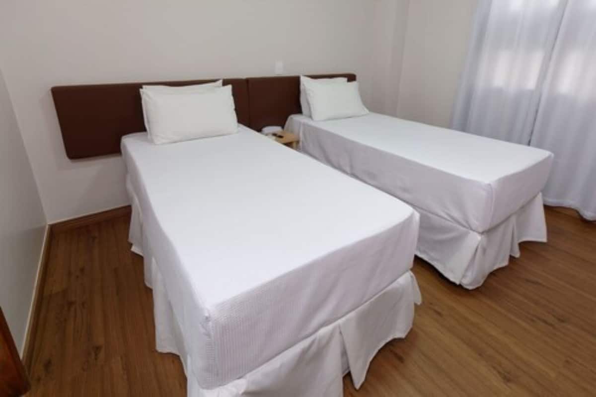 Quarto de hotel com duas camas de solteiro, piso de madeira e cabeceira branca simples.