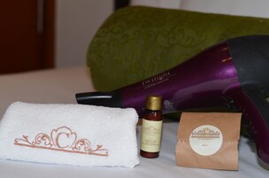Uma cama com travesseiro verde, um secador roxo, um frasco de shampoo La Concordia Suites, uma toalha branca com flores bordadas e um saco de papel marrom com o logotipo do hotel.