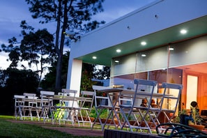 Uma área de estar externa com cadeiras e mesas dobráveis brancas, um edifício moderno com janelas amplas e um gramado bem cuidado.