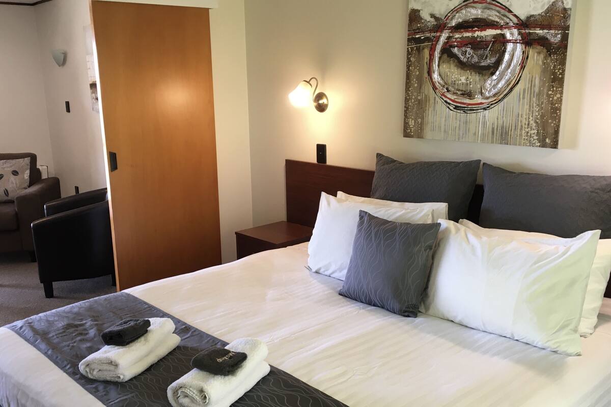Quarto de hotel com uma cama grande, cabeceira de madeira, mesa de cabeceira, luminária fixada na parede e um quadro.