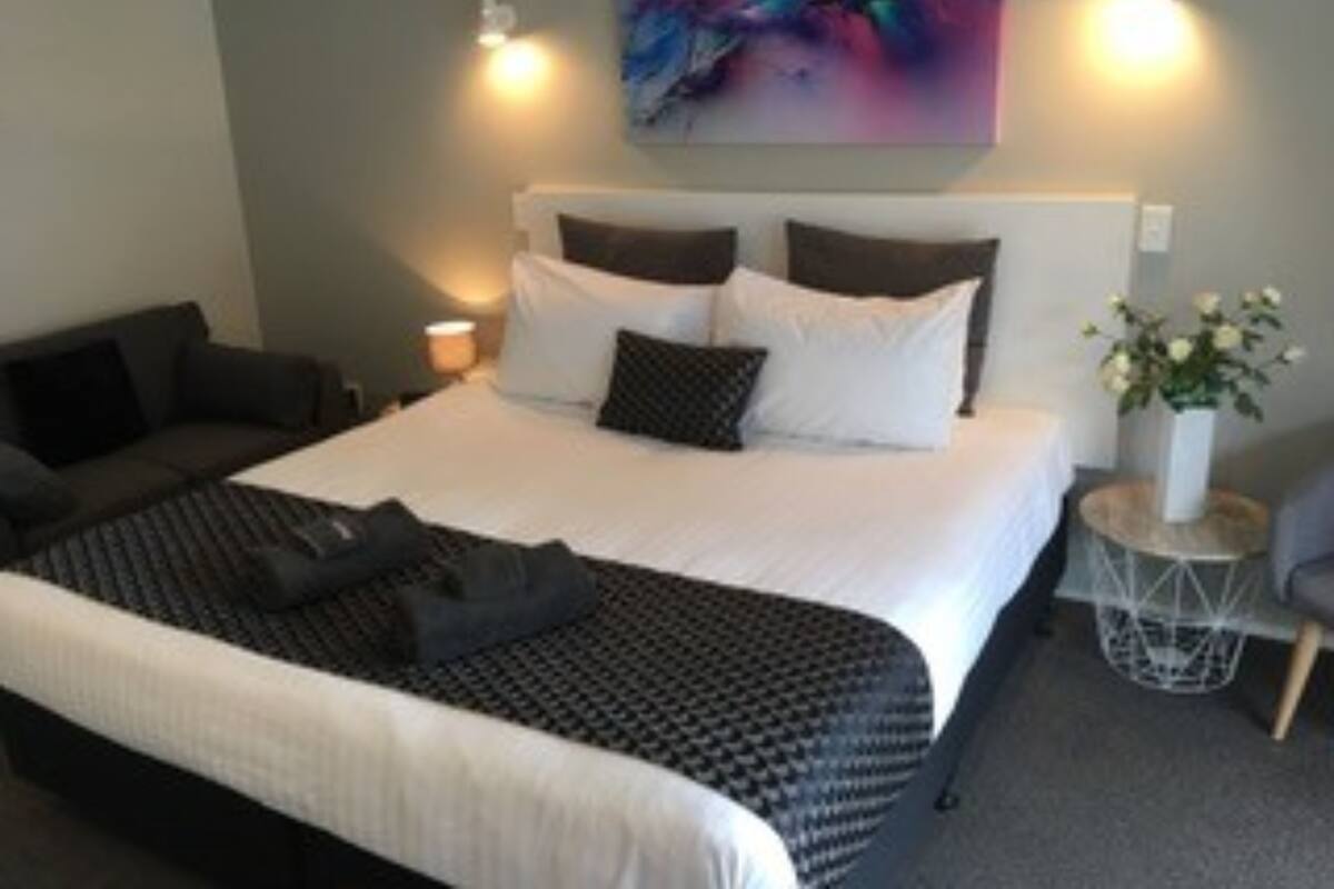 Quarto de hotel moderno com uma cama grande, uma mesinha de cabeceira, uma cadeira e um quadro na parede.