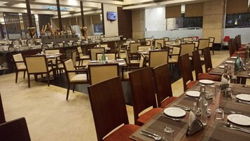 Uma ampla sala de jantar com várias mesas e cadeiras, área de cozinha com utensílios e uma tela de televisão.