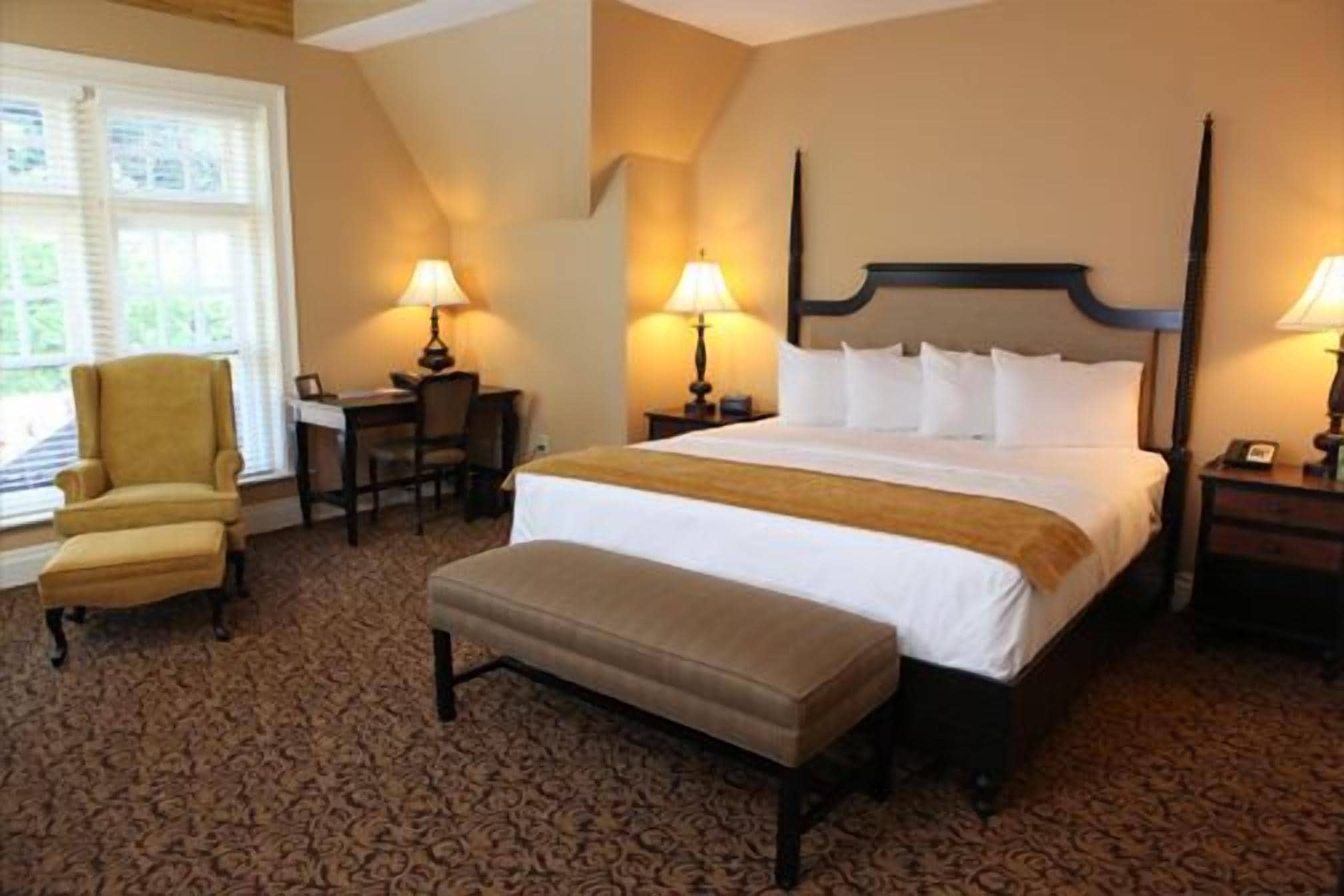 Quarto de hotel com cama de dossel, escrivaninha com cadeira, mesinha com abajur e um banquinho.