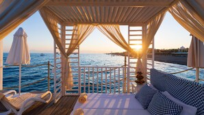 Uma cabana à beira-mar com um alpendre de madeira, assentos acolchoados brancos e vista para o oceano e o cais.