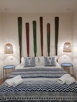 Cama bem arrumada com roupa de cama estampada, duas mesinhas de cabeceira e luzes decorativas fixadas na parede.