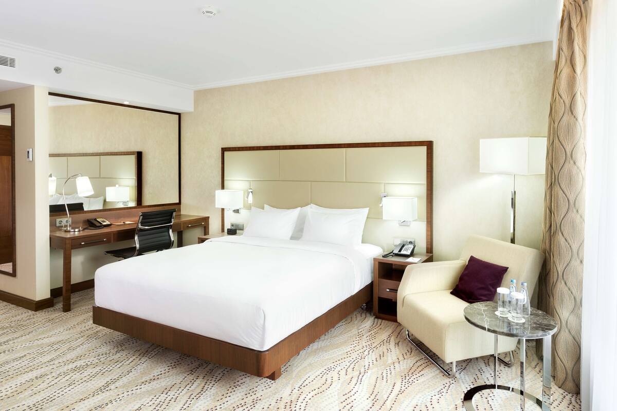 Quarto de hotel com uma cama grande, uma escrivaninha, uma cadeira, uma mesinha com garrafas e um espelho.