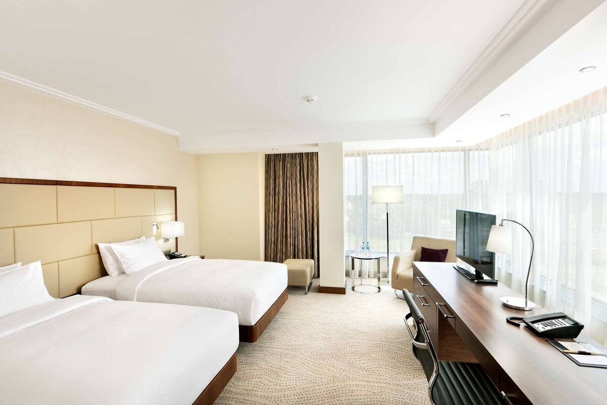Quarto de hotel com duas camas, uma mesa com computador, uma cadeira, televisão e janela com cortinas.