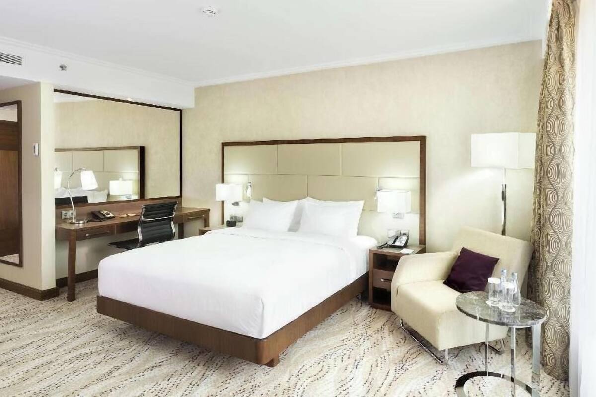 Quarto de hotel com uma cama grande, uma escrivaninha, uma cadeira, um sofá e uma mesinha com luminárias.