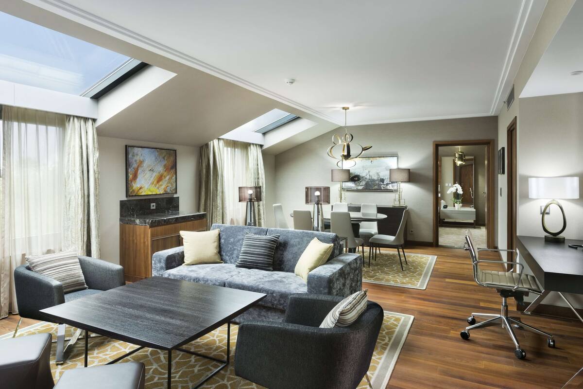 Quarto de hotel moderno com sala de estar, sala de jantar e quarto. Apresenta um claraboia, uma pintura grande e uma escrivaninha com abajur.