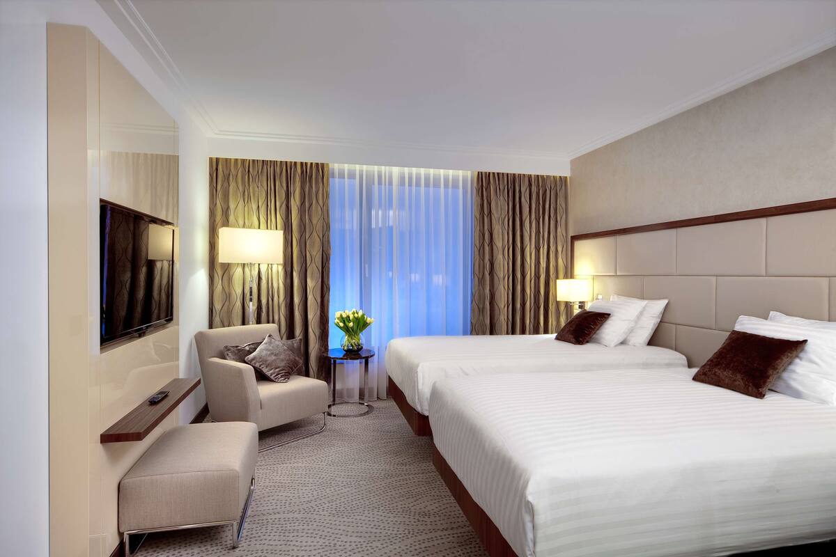 Quarto de hotel com uma cama grande, um sofá, uma TV e uma mesa pequena com um abajur e flores.