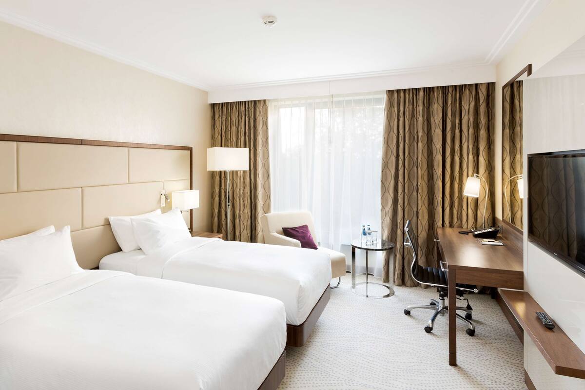 Quarto de hotel com duas camas, uma escrivaninha, uma cadeira, uma televisão e uma janela com cortinas.