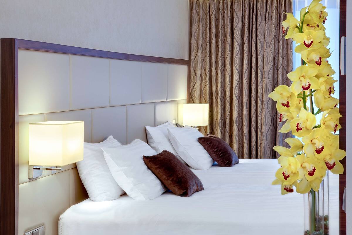Quarto de hotel com uma cama grande, duas luminárias de cabeceira, uma cortina decorativa e um vaso de flores amarelas.