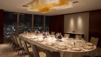 Uma sala de jantar com uma mesa comprida posta para uma refeição, grandes janelas e vista noturna da cidade.