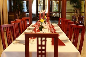 Uma longa mesa de jantar posta com guardanapos vermelhos, toalha de mesa branca e copos, ladeada por cadeiras de madeira num espaço com grandes janelas e plantas em vasos.