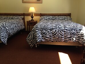 Quarto de hotel com duas camas, cada uma com roupa de cama estampada em zebra, cabeceira de madeira, mesa de cabeceira com luminária e piso coberto por carpete.