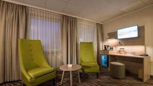 Quarto de hotel com duas poltronas verdes, uma mesa redonda pequena, uma escrivaninha com um computador e uma televisão exibindo o nome do hotel.