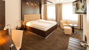 Quarto de hotel moderno com uma cama grande, área de estar com uma poltrona e uma mesinha, TV fixada na parede e uma janela ampla com cortinas.